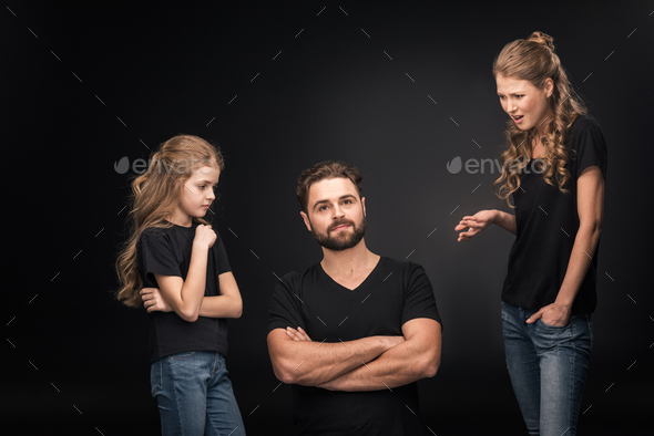 Mother And Daughter Quarreling At Father Sitting With Crossed Arms
