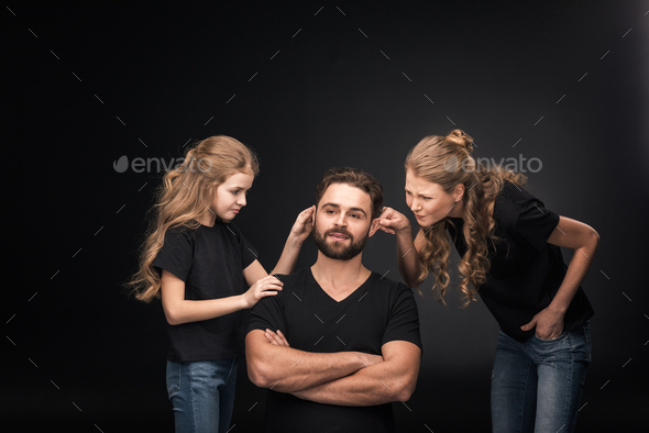 Mother And Daughter Quarreling At Father Sitting With Crossed Arms