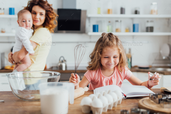 selective focus of child holding whisk and reading cookbook near table ...