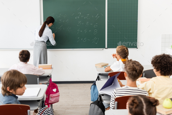 back view of teacher writing equations on chalkboard near multicultural ...