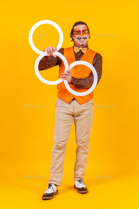 Happy juggler man in makeup vest juggling hoops on a yellow background ...