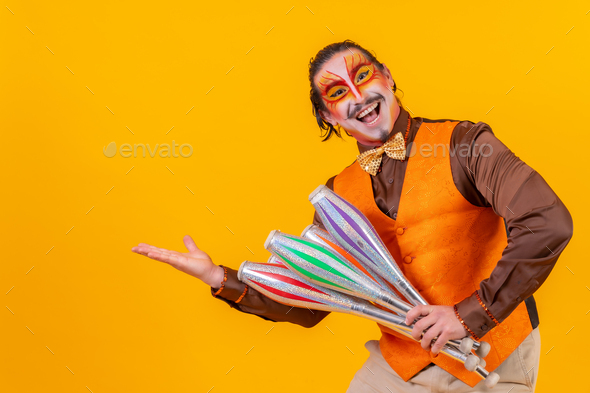 Happy juggler man in makeup vest juggling maces on a yellow background ...