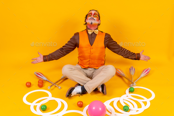 Juggler in a vest and with his face painted sitting with the juggling objects on a yellow ...