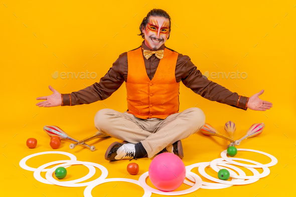 Juggler in a vest and with his face painted sitting with the juggling ...