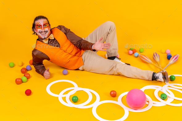 Juggler in a vest and with a painted face lying with the juggling objects on a yellow background ...