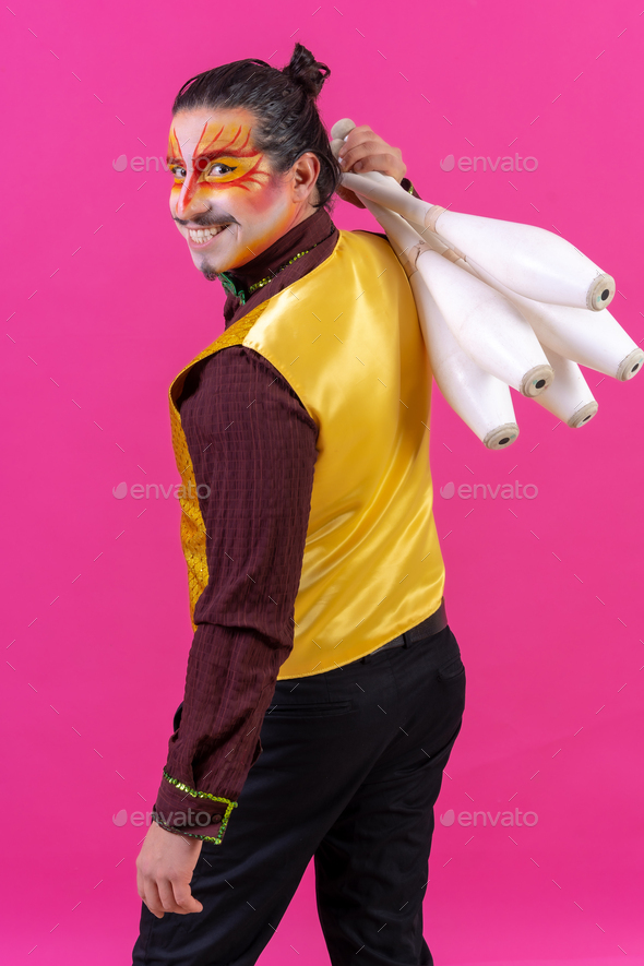 Portrait of a juggler in a waistcoat and with painted face juggling maces on a pink background ...