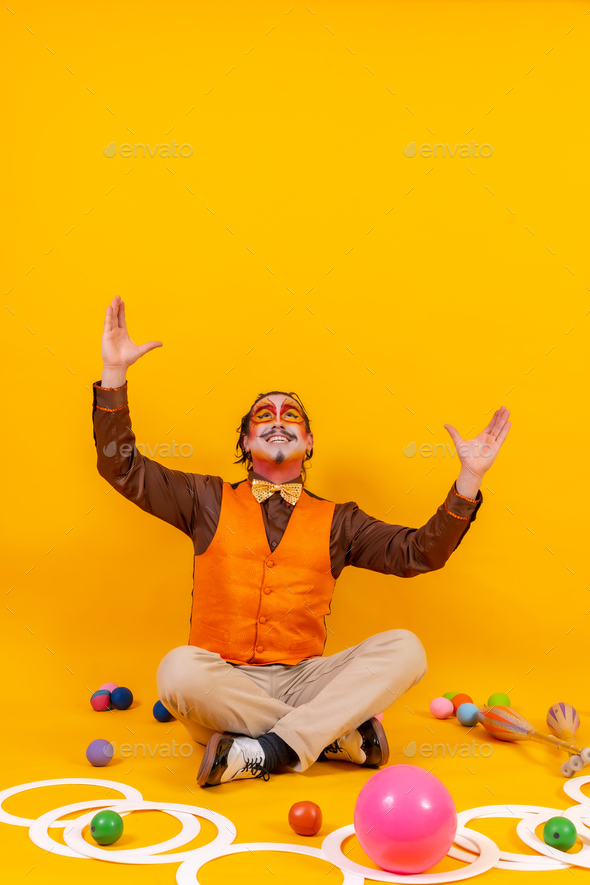 Juggler in a vest and with his face painted sitting with the juggling objects on a yellow ...