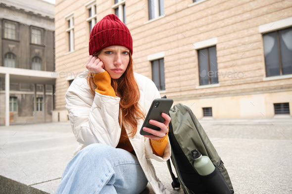 Redhead 20s years girl, sits with smartphone outside building, looks complicated and upset ...