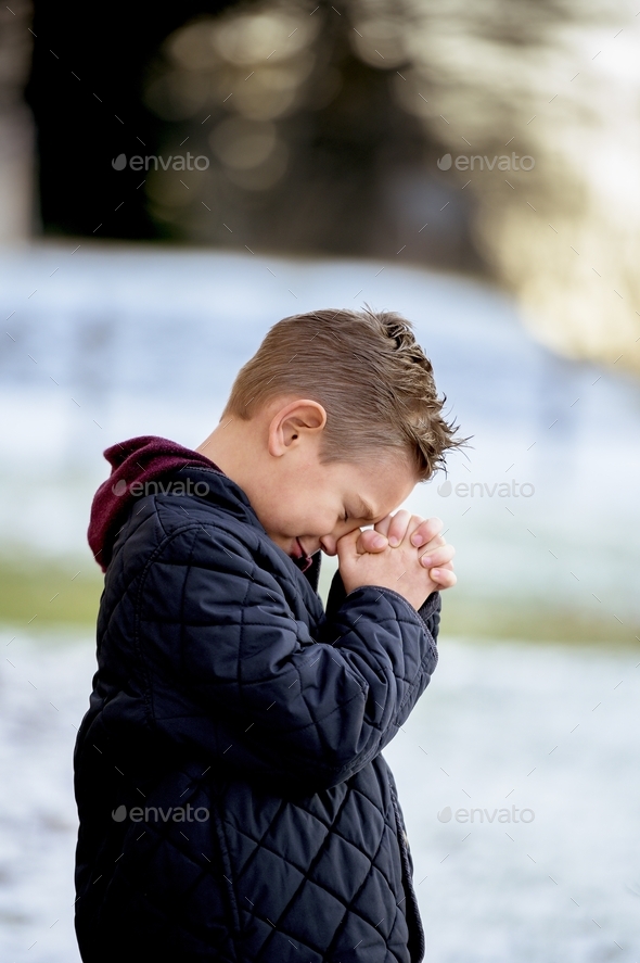 Little boy standing in a park and praying under sunlight with a blurry ...