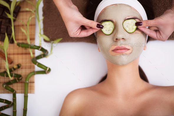 Spa. Beautiful young woman is getting facial clay mask at spa, lying with cucumbers on eyes ...