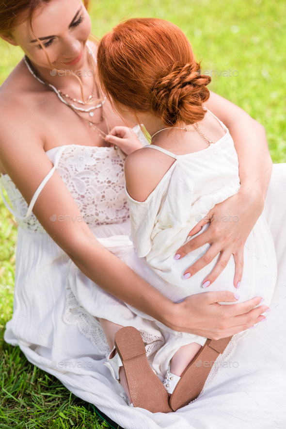 beautiful young mother with cute redhead daughter wearing white dresses and hugging at park ...