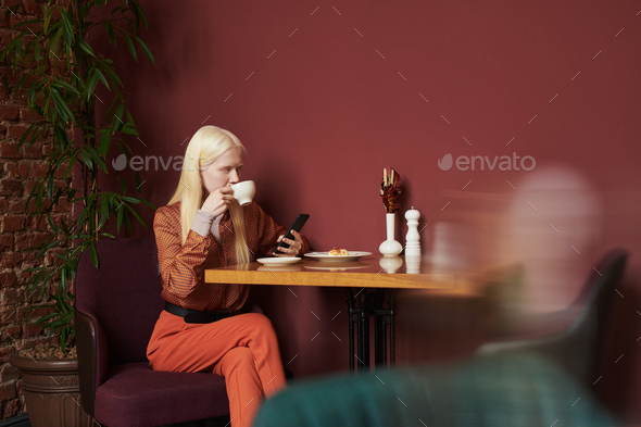 Restful albino girl in smart casualwear having coffee or tea with ...