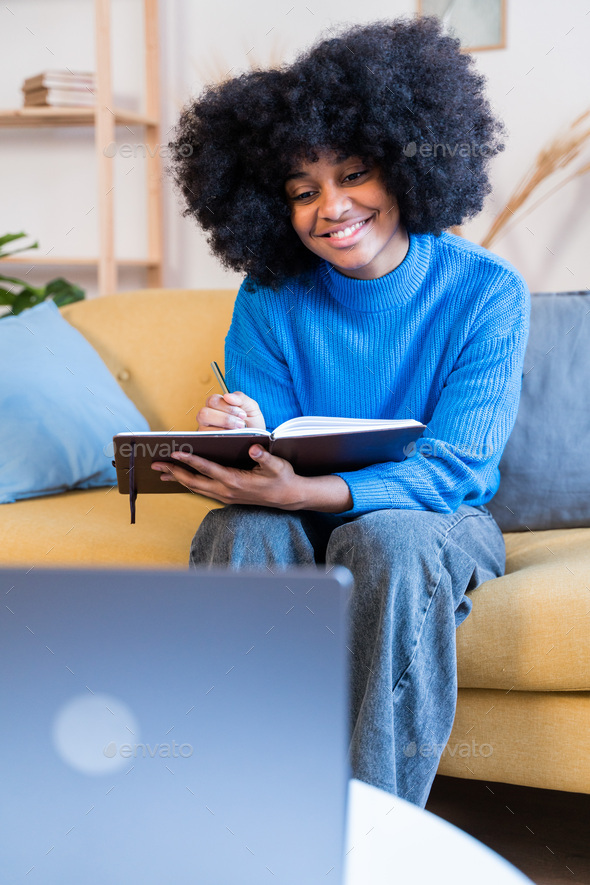 Smiling black woman taking notes while attending a university online ...
