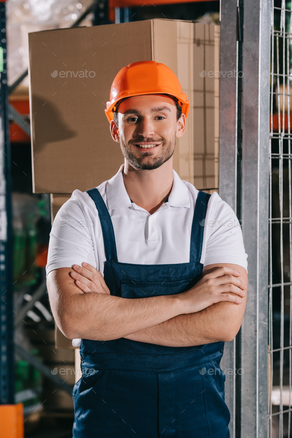 handsome warehouse worker smiling at camera while standing with crossed ...