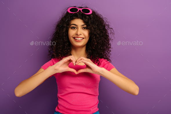 happy bi-racial girl showing heart sign with hands while looking at ...