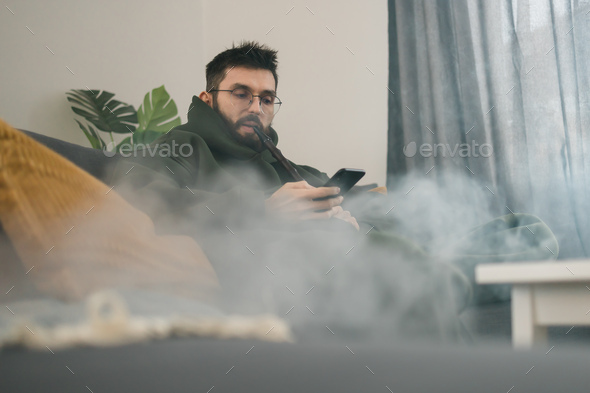 Bearded millennial or gen z man smoking hookah while relaxing on sofa ...