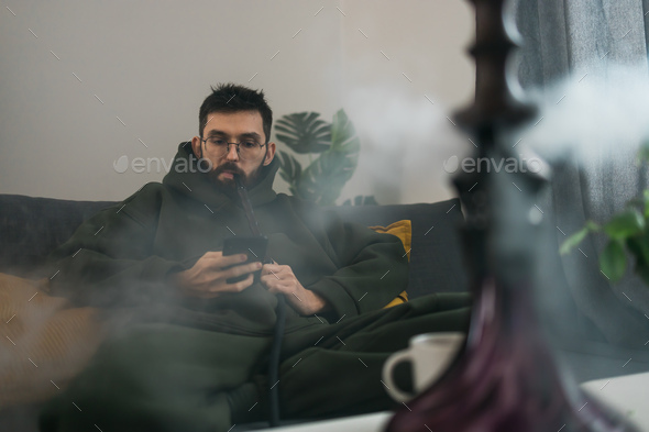 Bearded millennial or gen z man smoking hookah while relaxing on sofa ...
