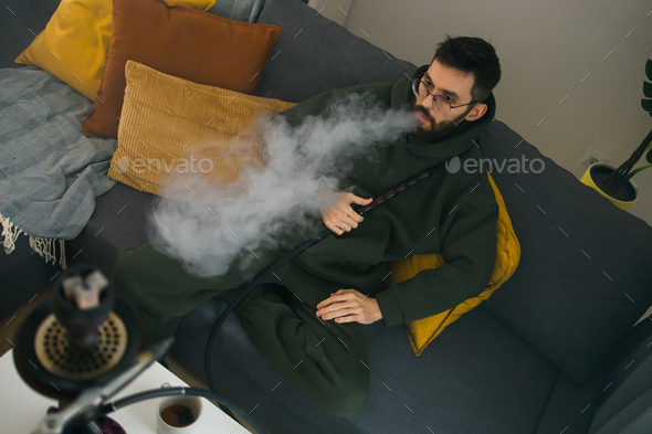 Bearded millennial or gen z man smoking hookah while relaxing on sofa ...
