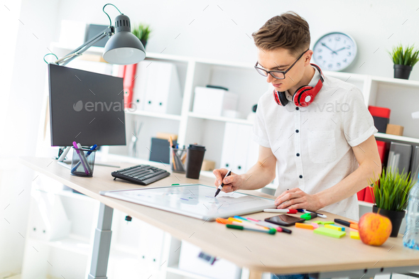 A young man in glasses stands near a computer desk. A young man draws a ...