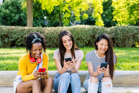 Teenage woman friends using mobile phone together sitting outdoors ...