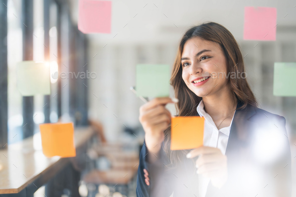 Young smiley attractive, businesswoman using sticky notes in glass wall ...