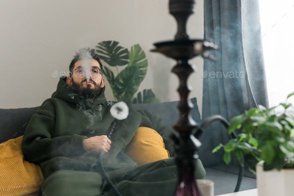 Bearded millennial or gen z man smoking hookah while relaxing on sofa ...
