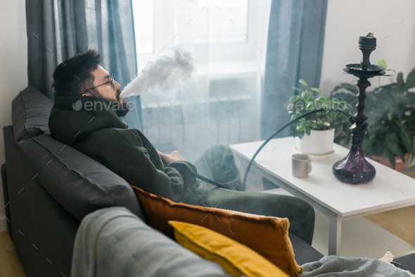 Bearded millennial or gen z man smoking hookah while relaxing on sofa ...