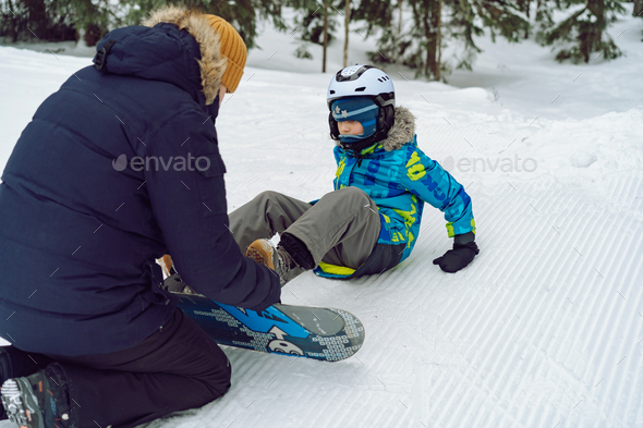 father helping little boy sitting on snow putting his feet in snowboard ...