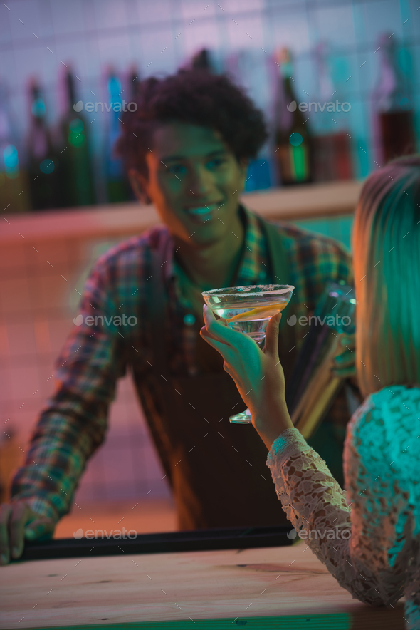 back view of woman with cocktail having conversation with barman in bar ...