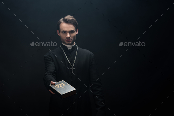 young serious catholic priest holding holy bible in outstretched hand ...