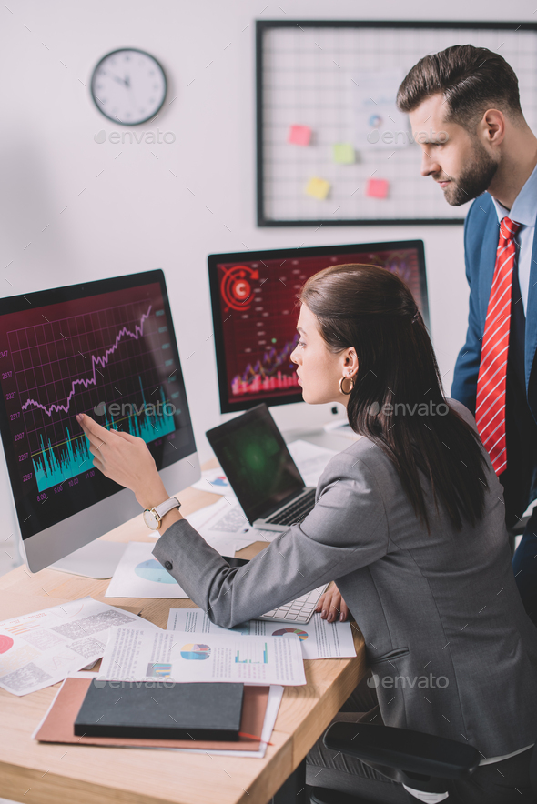 Side view of data analyst pointing with hand at graphs on computer monitor to colleague in ...