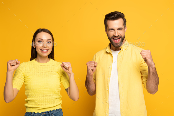 Cheerful couple showing yeah gesture at camera on yellow background ...