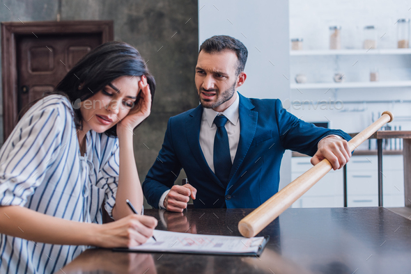 Scared woman writing in documents near angry collector with baseball ...