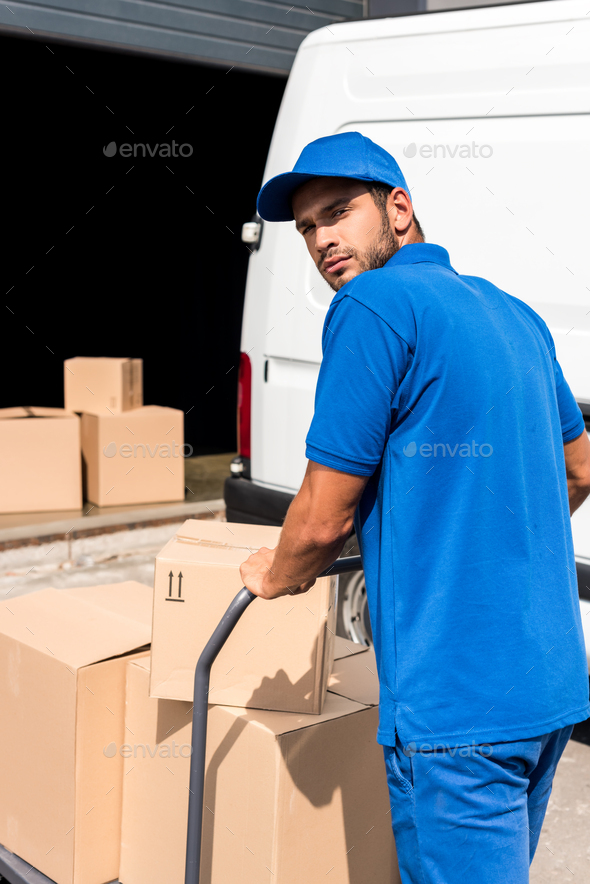 serious delivery man driving cart with cardboard boxes into warehouse ...