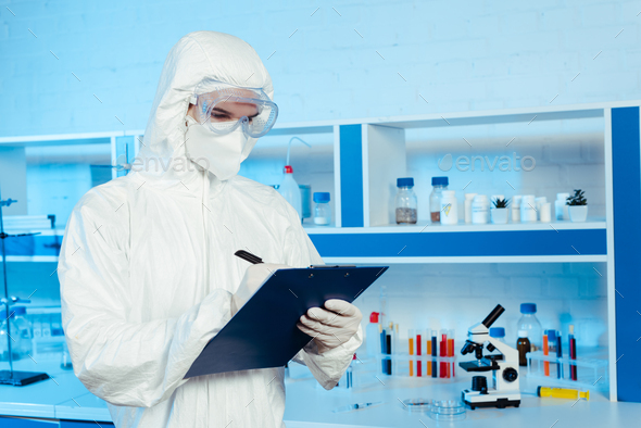 scientist in hazmat suit and goggles writing while holding clipboard ...