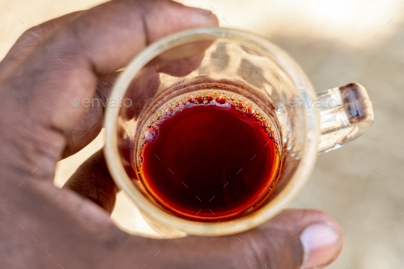Cup of red raw tea held in fingers. Red tea and its bubbles are seen ...