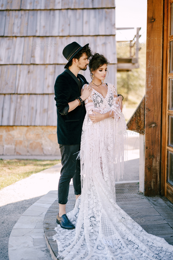Groom hugs bride in a lace braided dress on the doorstep of a wooden ...