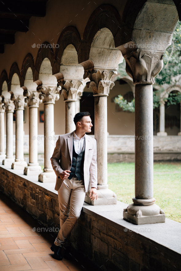 Groom in a suit stands leaning on the windowsill of a loggia with ...