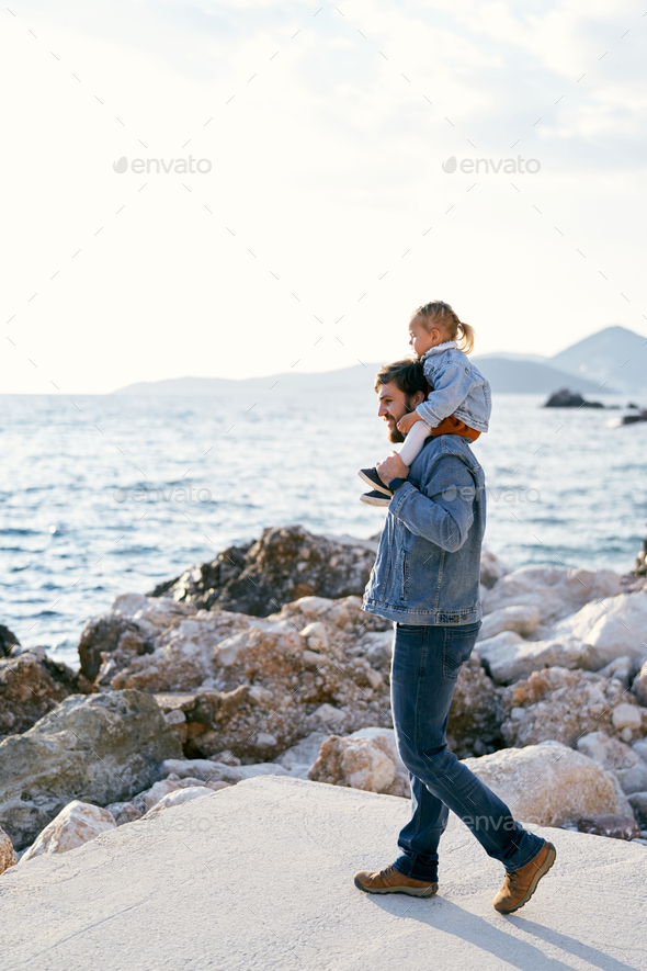 Smiling dad carries a little girl on his shoulders while walking along the pebble beach to the ...