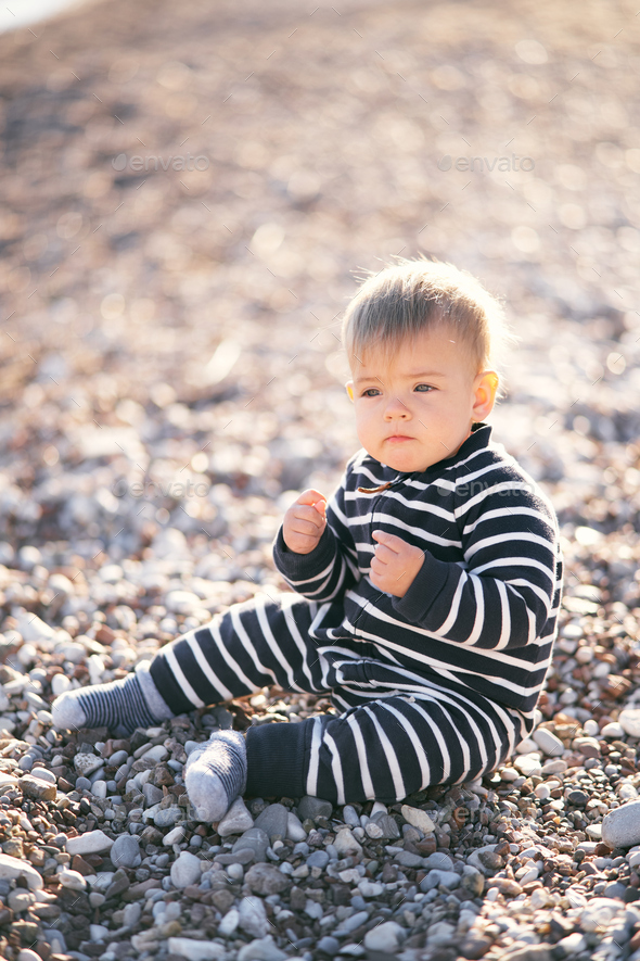 Serious toddler in striped overalls sits on a pebble beach, holding his ...