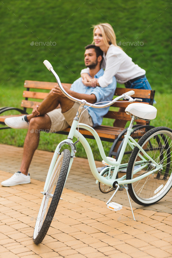 selective focus of bicycle and young couple resting on bench in park ...