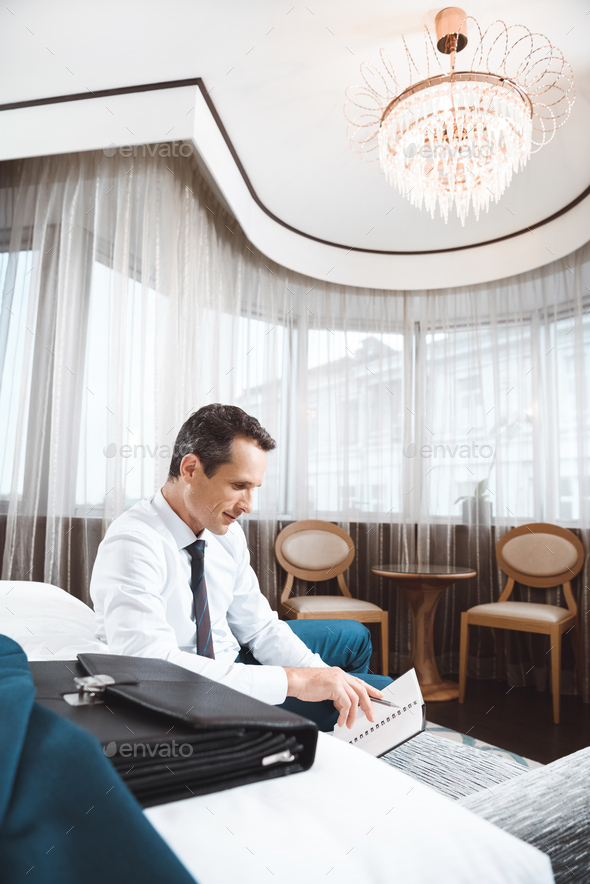 Businessman in formal wear sitting on bed in hotel room and reading ...