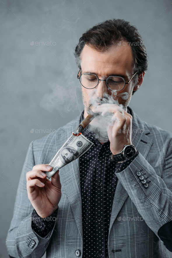 confident businessman in eyeglasses smoking cigar and burning dollar ...