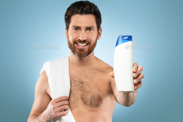 Happy handsome man showing shampoo bottle, standing with towel over ...