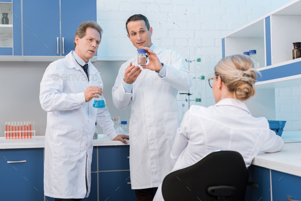 Two professional scientists showing their colleague jars with chemicals ...
