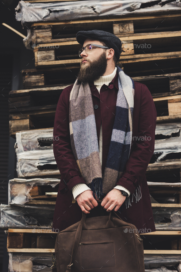 handsome bearded stylish man with leather bag in front of industrial ...
