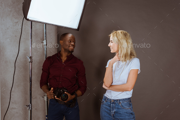 cheerful multiethnic photographer and model looking at each other while ...