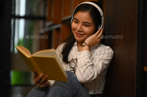 Pleasant young woman listening to music on headphone and reading book ...