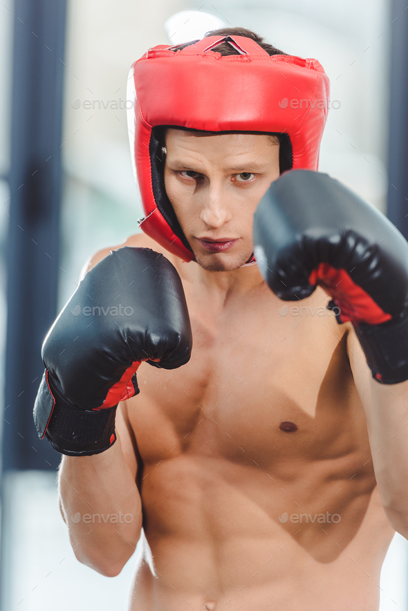 young shirtless muscular boxer looking at camera while boxing in gym ...