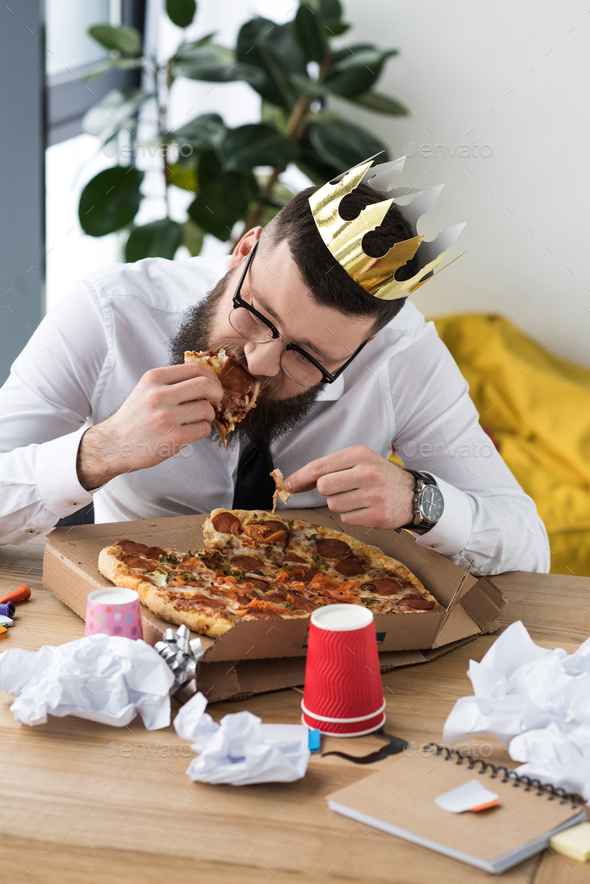 portrait of businessman with paper crown on head eating pizza at ...